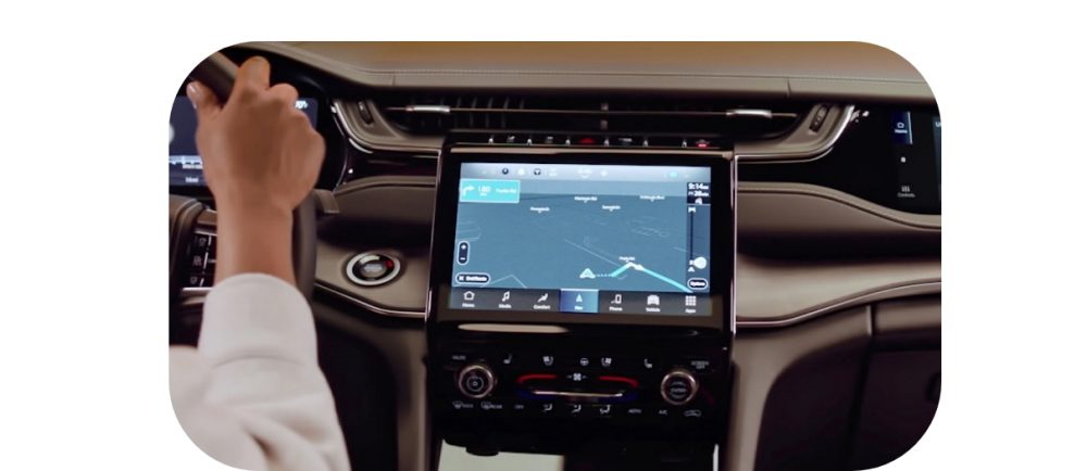 The interior of a 2024 Jeep Grand Cherokee with a woman driving and the Uconnect touchscreen ...